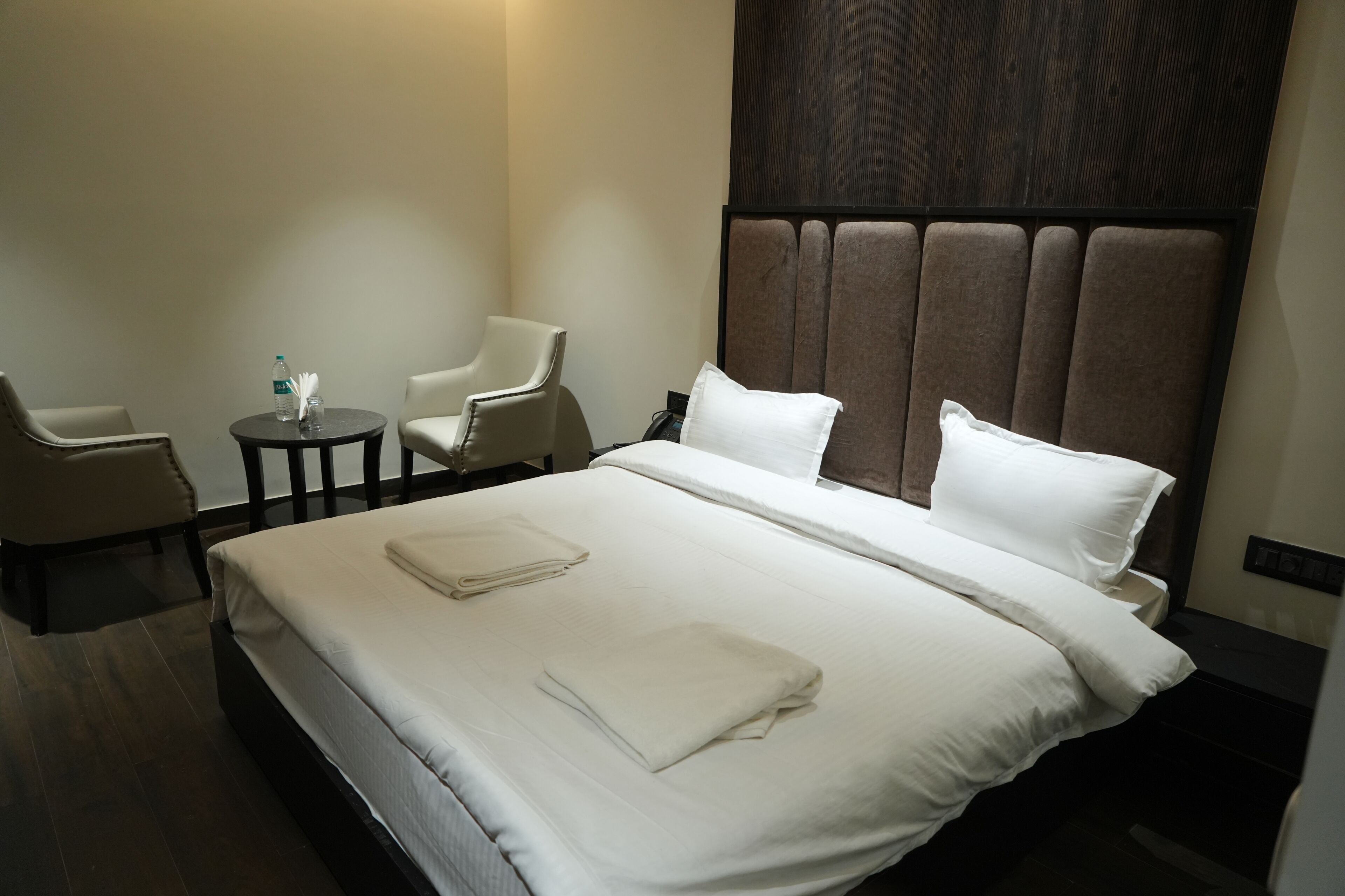 Quarto de hotel com uma cama grande, duas poltronas, uma mesinha redonda com uma garrafa e guardanapos, e uma cabeceira de madeira.