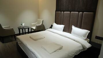 Quarto de hotel com uma cama grande, duas poltronas, uma mesinha redonda com uma garrafa e guardanapos, e uma cabeceira de madeira.