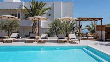 Uma área de piscina com espreguiçadeiras e guarda-sóis, uma piscina azul cristalina e um edifício branco ao fundo.