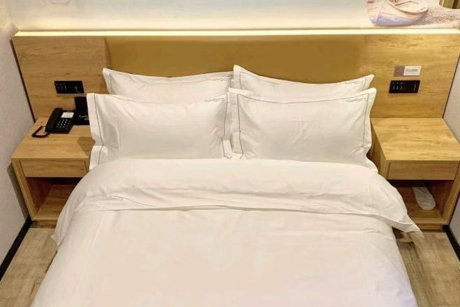 Uma cama bem arrumada com lençóis brancos, um telefone ao lado da cama e uma pequena mesa de cabeceira de madeira.