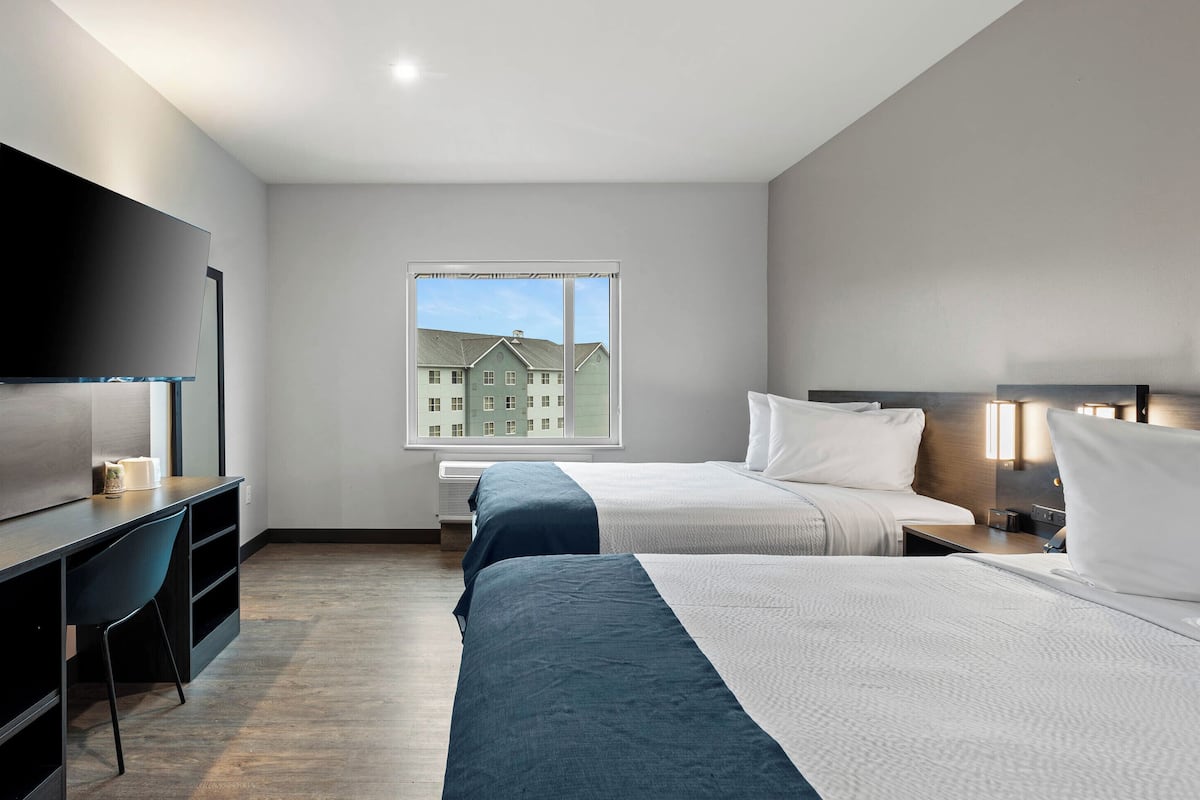 Quarto de hotel com escrivaninha, duas camas, televisão e janela com vista para edifícios.