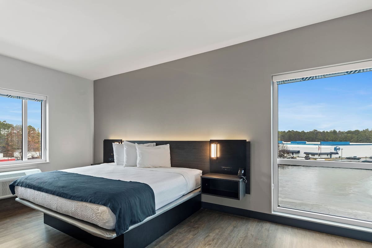 Quarto de hotel moderno com uma cama grande, mesa de cabeceira, luminária na parede e vista para um corpo d'água e edifícios industriais.