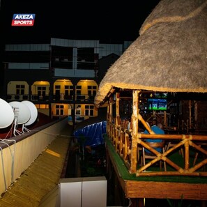 Estrutura com telhado de palha e guarda-corpo de madeira, provavelmente um bar ou área de lounge, em uma varanda com antenas parabólicas e vista para um edifício com várias janelas.