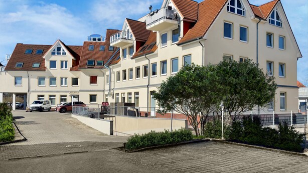 Uma fileira de edifícios modernos de vários andares, com telhados de telhas vermelhas, um caminho de entrada pavimentado e uma pequena área ajardinada com árvores.