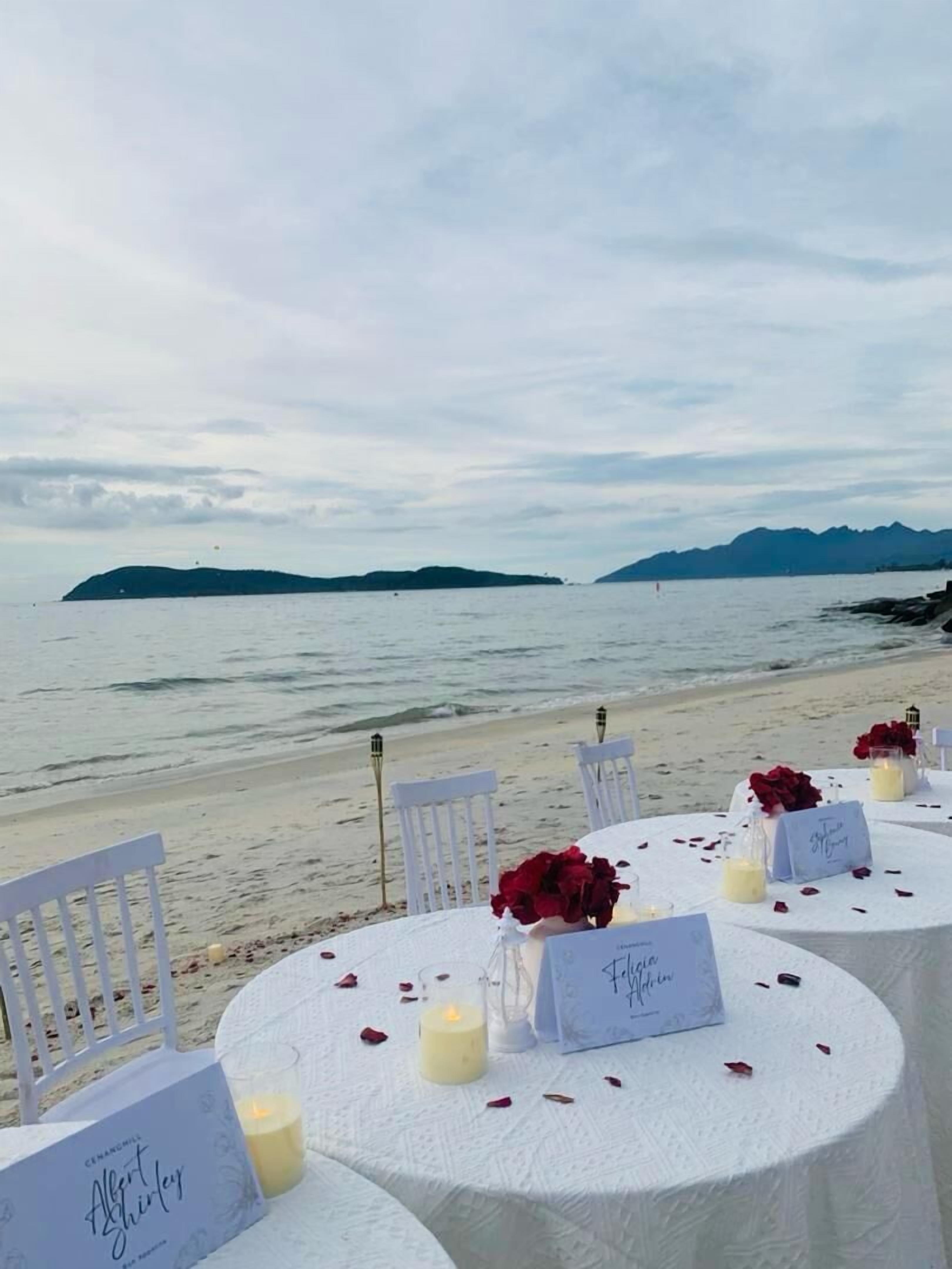 Mesas redondas com toalhas brancas, rosas vermelhas, velas e cartões de lugar em ambiente praiano.