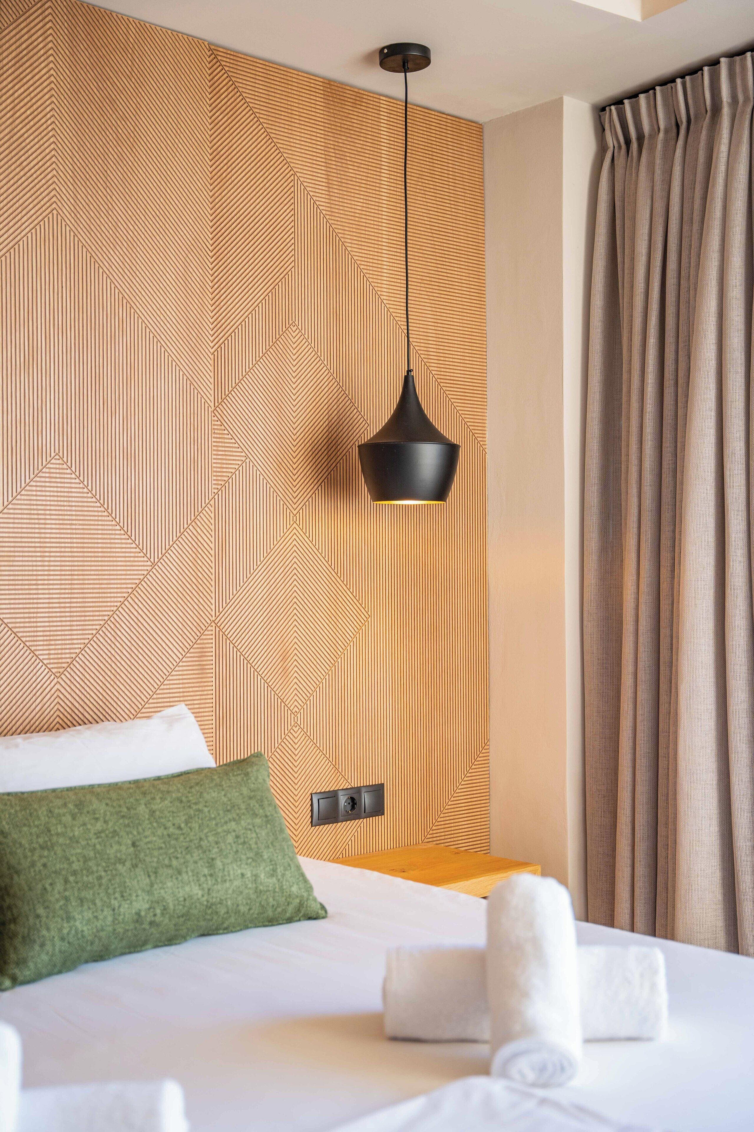 Quarto de hotel com parede estampada em padrão geométrico, luminária pendente, cama com roupa de cama branca, travesseiro verde e um conjunto de toalhas brancas.