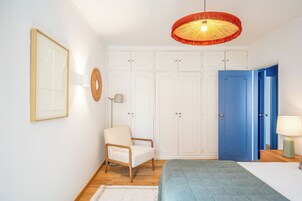 Quarto com cama, cadeira, guarda-roupa, porta azul e luminária suspensa.