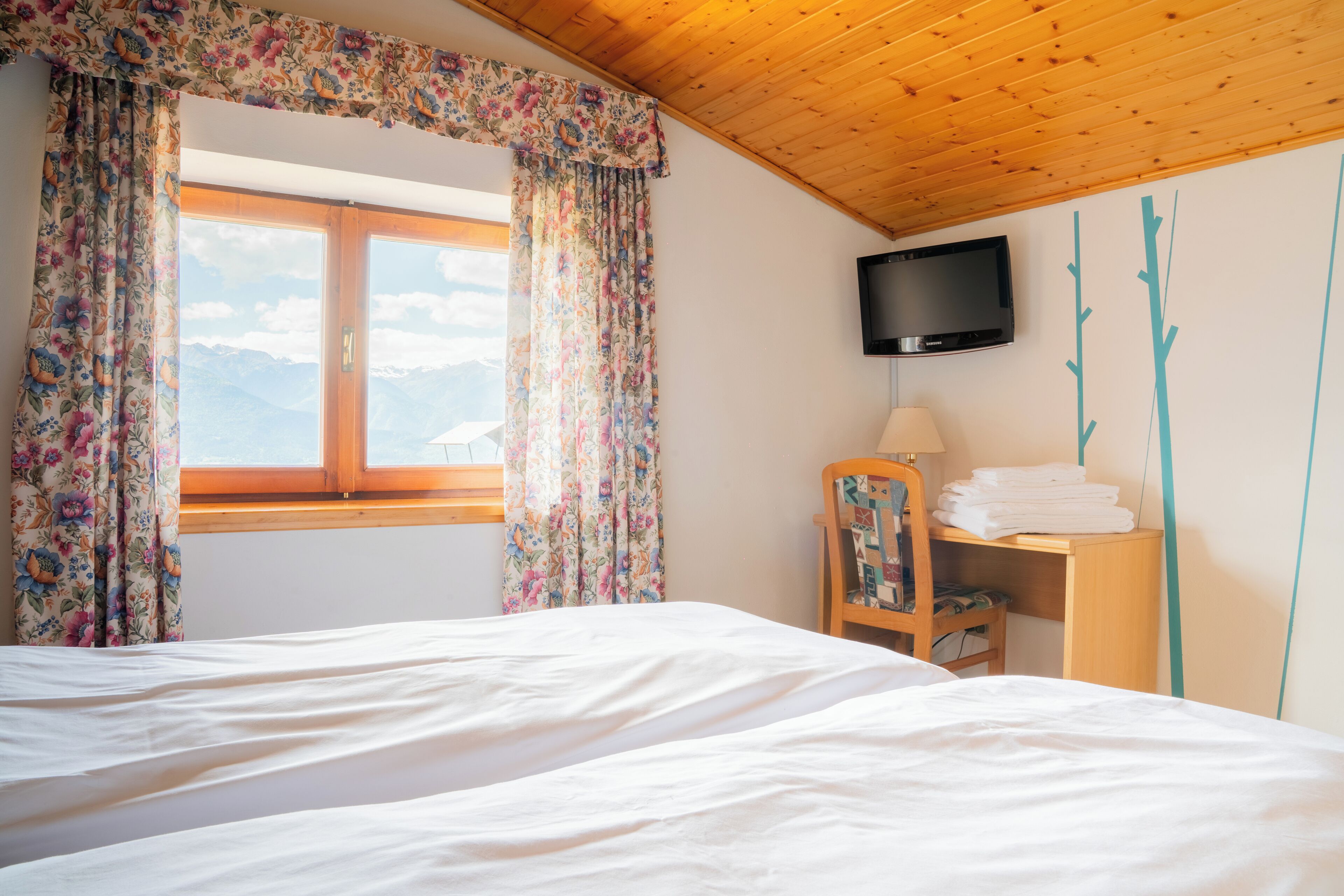 Quarto com teto de madeira, uma cama, televisão na parede e janela com cortinas.