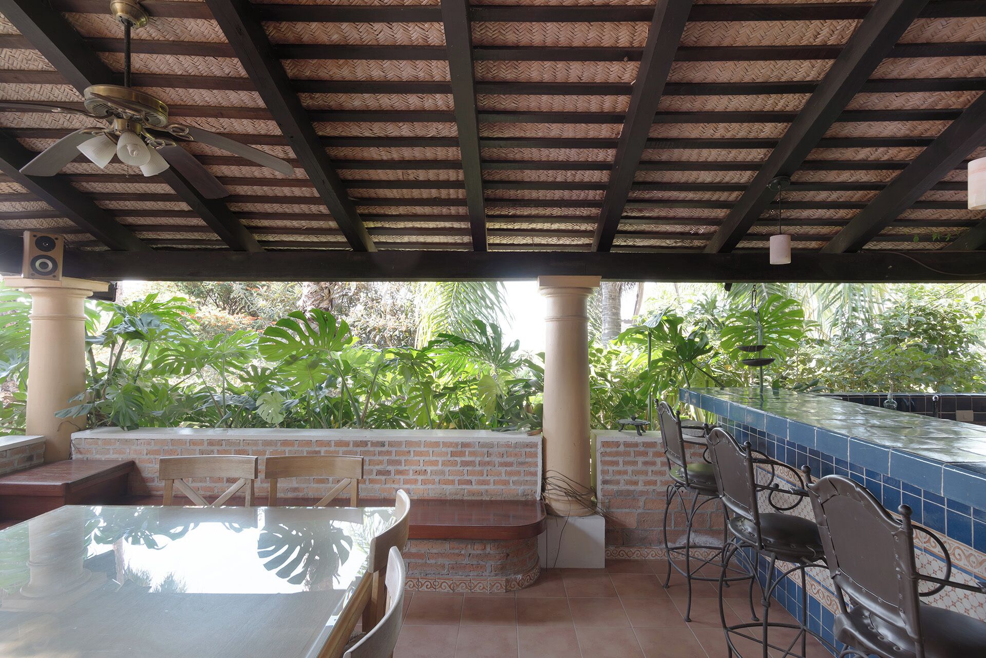 Área externa coberta com mesa, cadeiras e um ventilador de teto, com vista para vegetação exuberante.