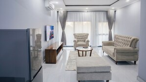 Uma sala de estar moderna com sofá, poltrona e mesa de centro, uma TV de tela plana e uma janela grande com cortinas.