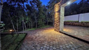 Um espaço de pátio pavimentado com azulejos hexagonais, uma parede de pedra com luzes embutidas e uma área arborizada ao redor.