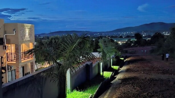 Um edifício com várias andares e varandas, vista para a cidade ao entardecer, e um caminho que leva a uma área gramada.
