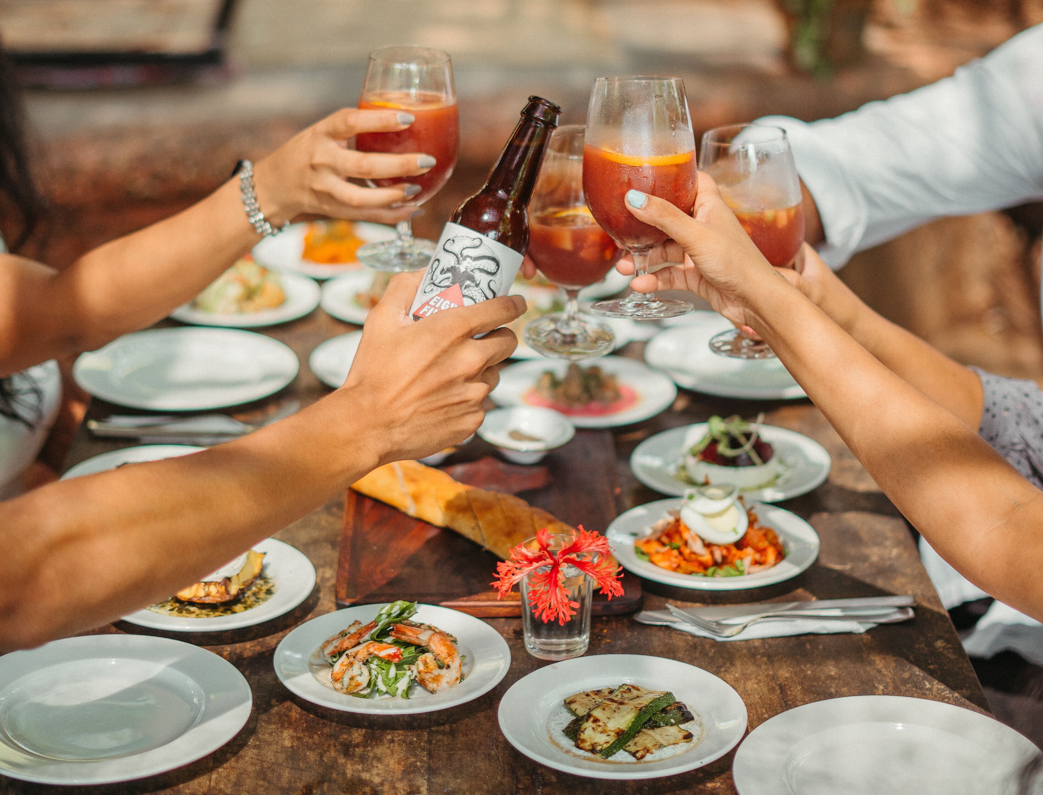 Um grupo de pessoas brindando com bebidas em uma mesa de madeira, com diversos pratos.