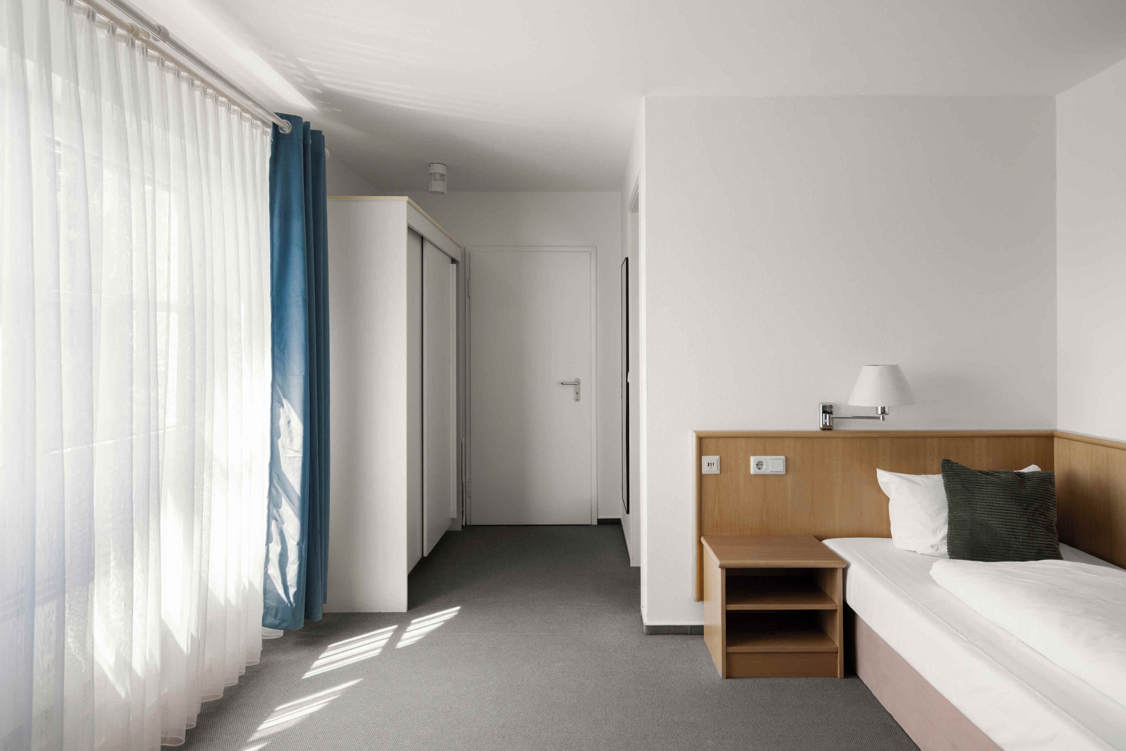 Quarto de hotel com uma cama, mesa de cabeceira, luminária e vista para um corredor.