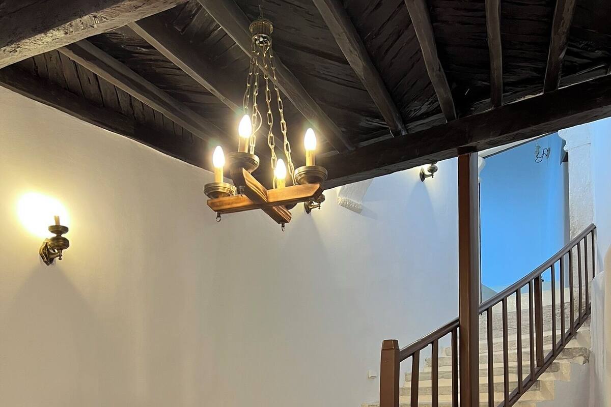 Uma escadaria com degraus de pedra e corrimão de madeira, conduzindo a um quarto com parede branca e um candelabro pendurado.