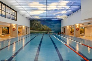 Uma grande piscina coberta com faixas divisórias bem definidas, um mural com paisagem tropical no teto e boias salva-vidas nas paredes.