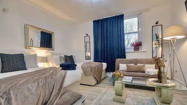 Quarto de hotel moderno com cama, sofá, mesa de centro e uma grande janela com cortinas.