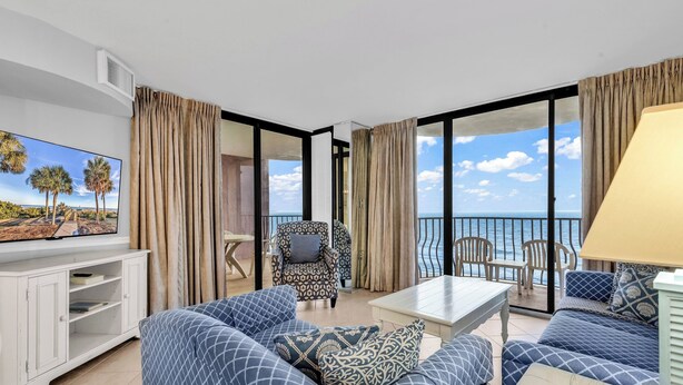 Sala de estar moderna com uma televisão grande, sofás estampados em azul, uma mesa de centro branca e vista para o mar através de grandes janelas.