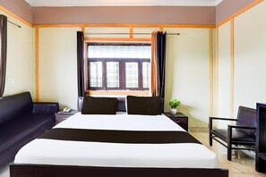 Quarto de hotel com uma cama grande, uma escrivaninha com telefone, uma cadeira, uma mesinha com uma planta e uma janela com cortinas.