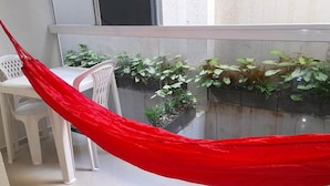 Uma rede vermelha, cadeiras de plástico branco e vasos com plantas numa varanda.