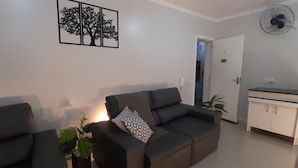 Uma sala de estar com sofá, uma mesinha com uma planta, um ventilador fixado na parede e uma porta que dá para outro cômodo.