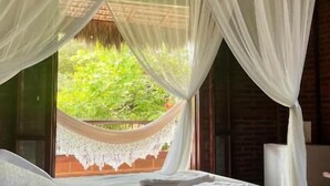 Quarto com cama, roupa de cama branca e vista para o verde através de cortinas leves.