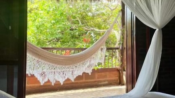 Quarto com cama, rede e vista para o verde através de uma janela com cortinas brancas.