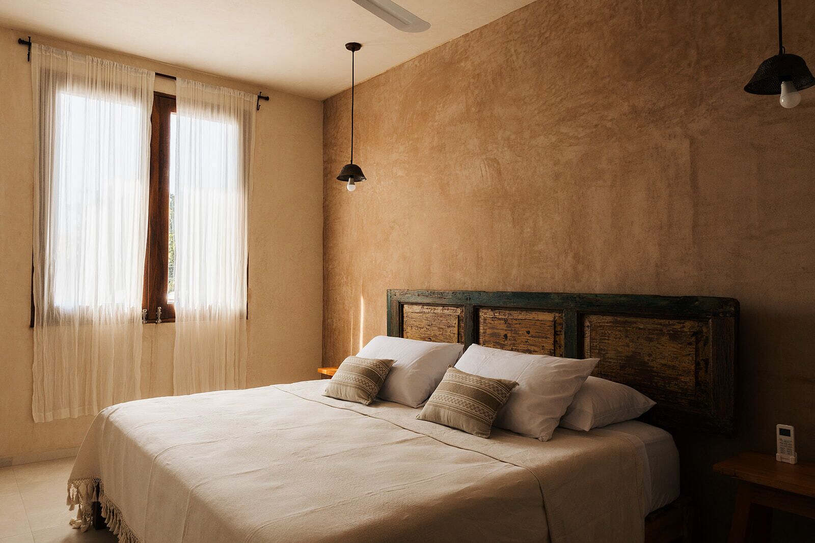 Quarto com cama, cabeceira de madeira, mesa de cabeceira, luminária e cortinas leves.