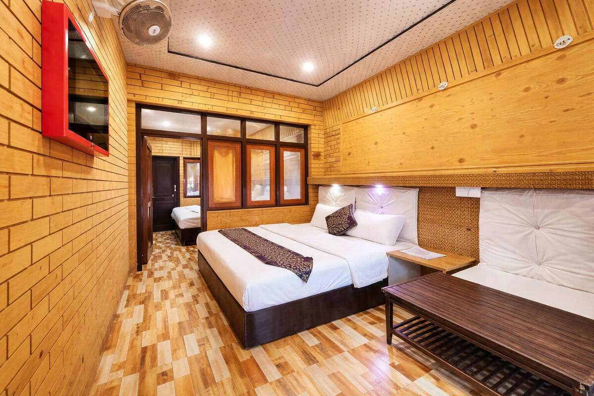 Quarto de hotel com piso de madeira, uma cama com roupa de cama branca, uma mesinha e um espelho com moldura vermelha.
