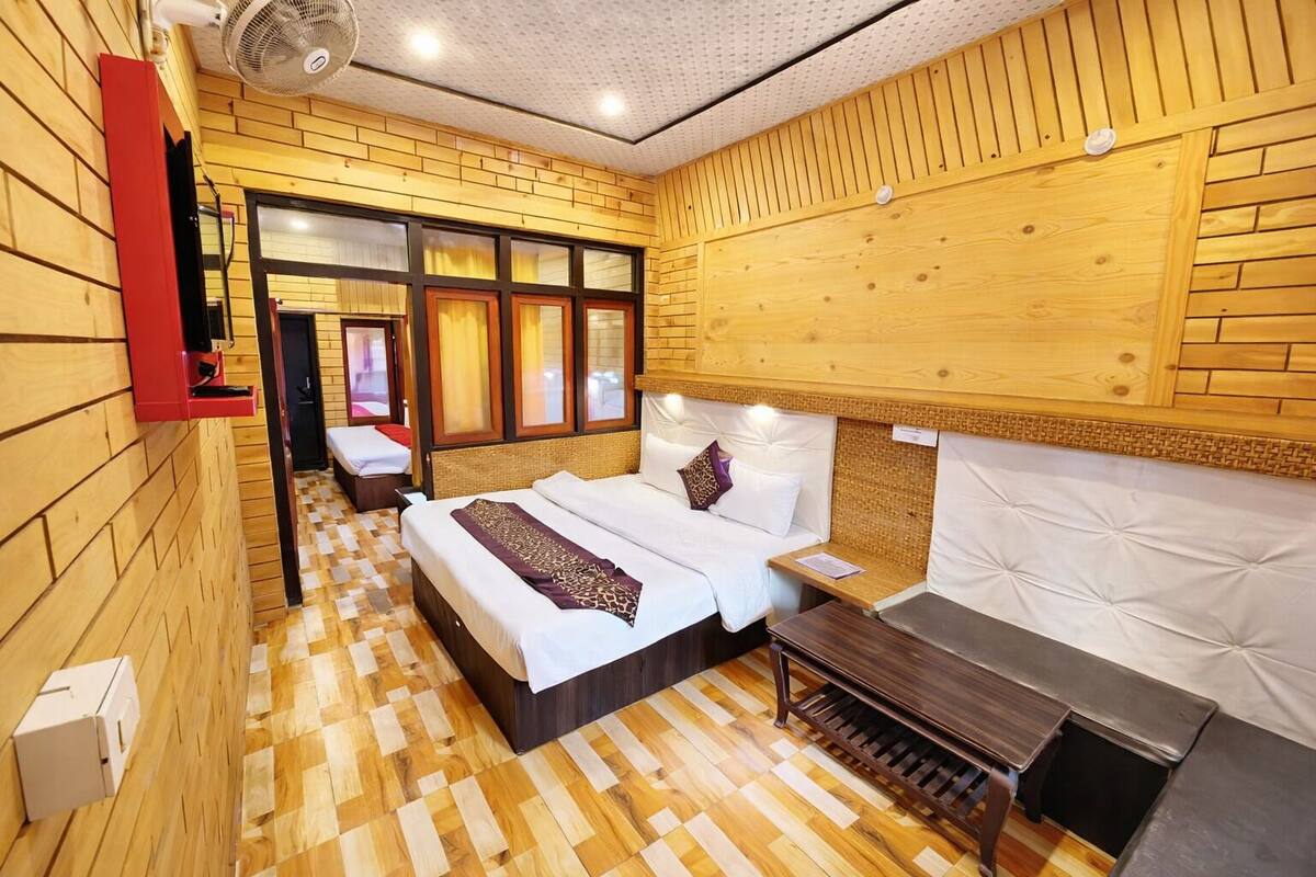 Quarto de hotel com piso de madeira, cama com roupa de cama branca, mesa de cabeceira e uma pequena escrivaninha.