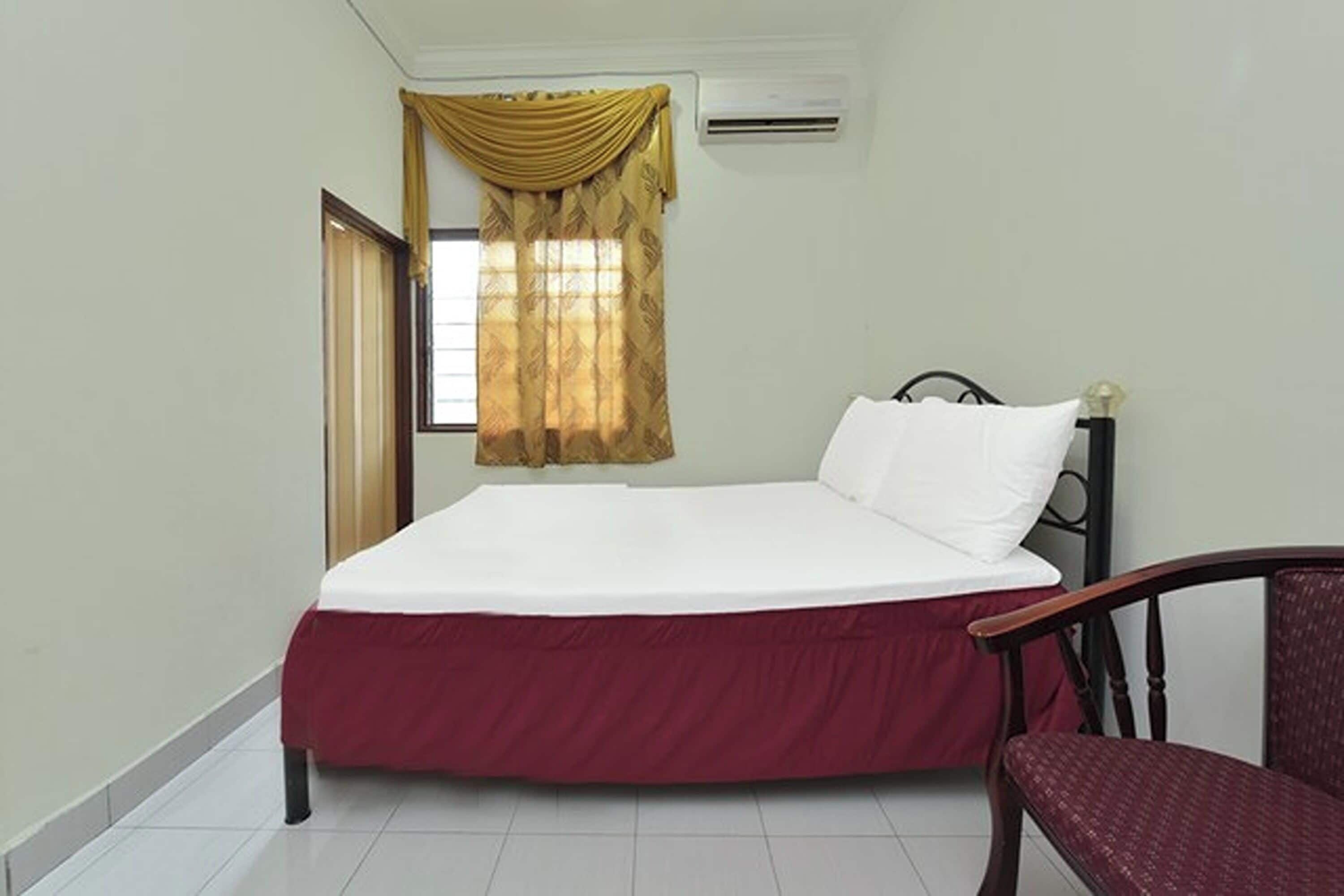 Uma cama de solteiro com colchão branco e edredom vermelho em um quarto com paredes brancas e uma janela com cortina amarela.