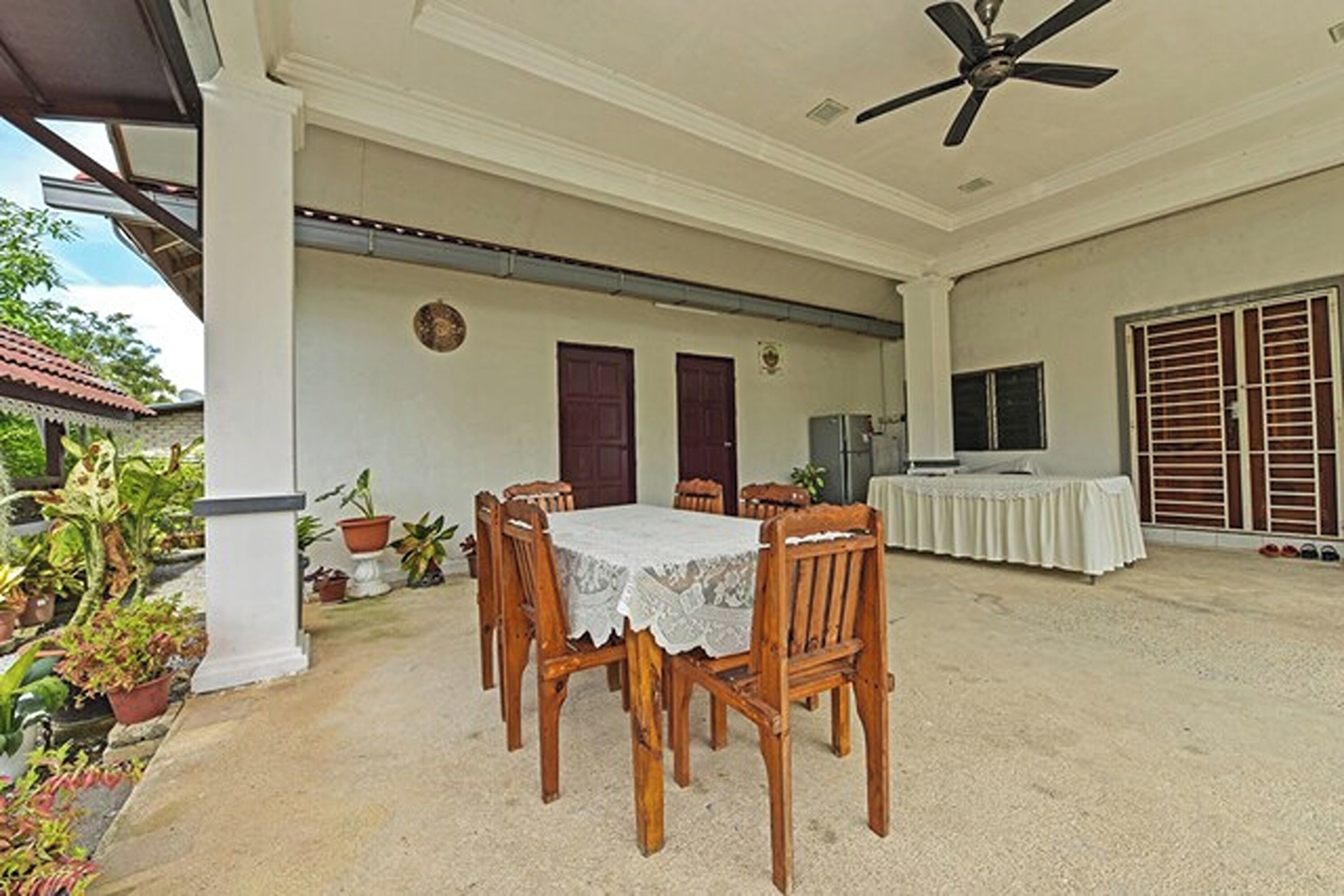 Um alpendre coberto com mesa, cadeiras, ventilador de teto e vasos de plantas.