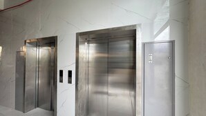 Três portas de elevador metálicas em um corredor com paredes de mármore e um cano vermelho no teto.