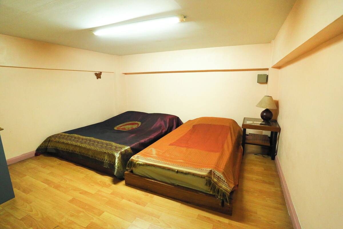 Quarto pequeno e simples com duas camas, mesa de cabeceira e luminária.