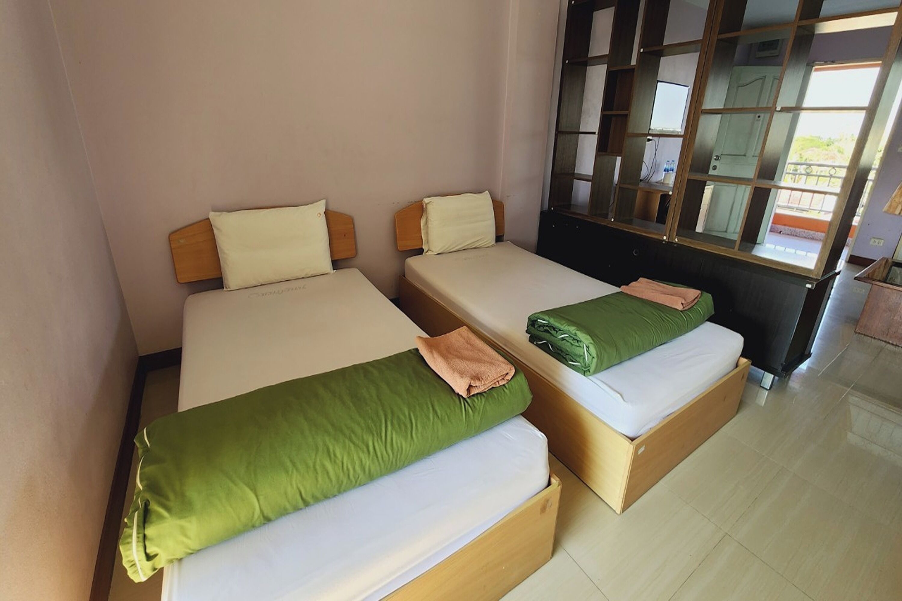Duas camas de solteiro com estruturas de madeira e roupa de cama branca, um cobertor verde em uma cama, e vista para o exterior por uma janela com grade de metal.