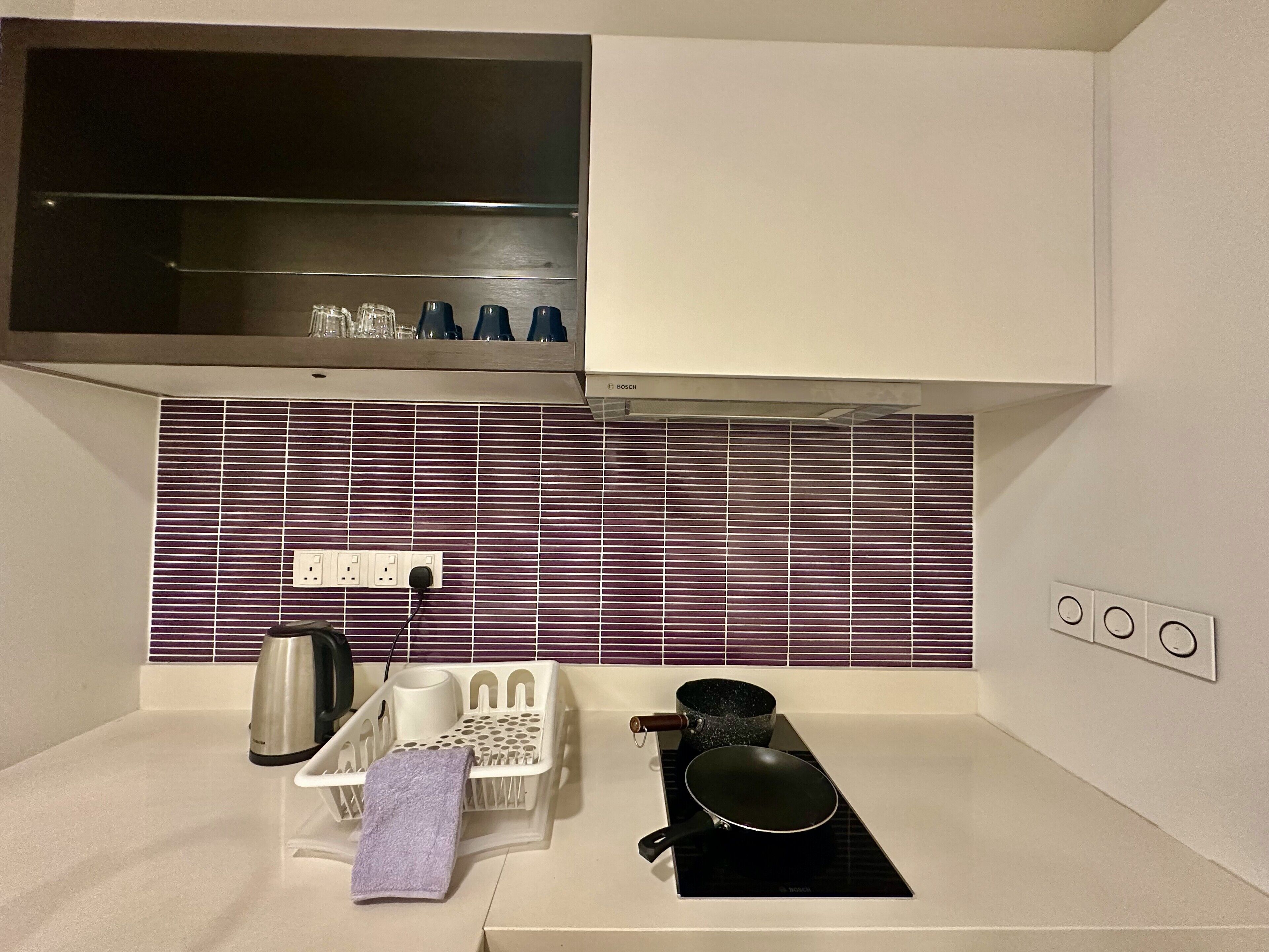 Uma cozinha com design moderno, apresentando um exaustor de aço inoxidável, painel de azulejos, chaleira, escorredor de louça e uma frigideira sobre uma placa de indução.