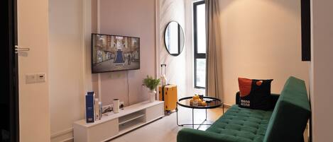 Uma sala de estar moderna com um sofá verde, uma TV de tela plana na parede, uma mesinha lateral e uma janela com cortinas.