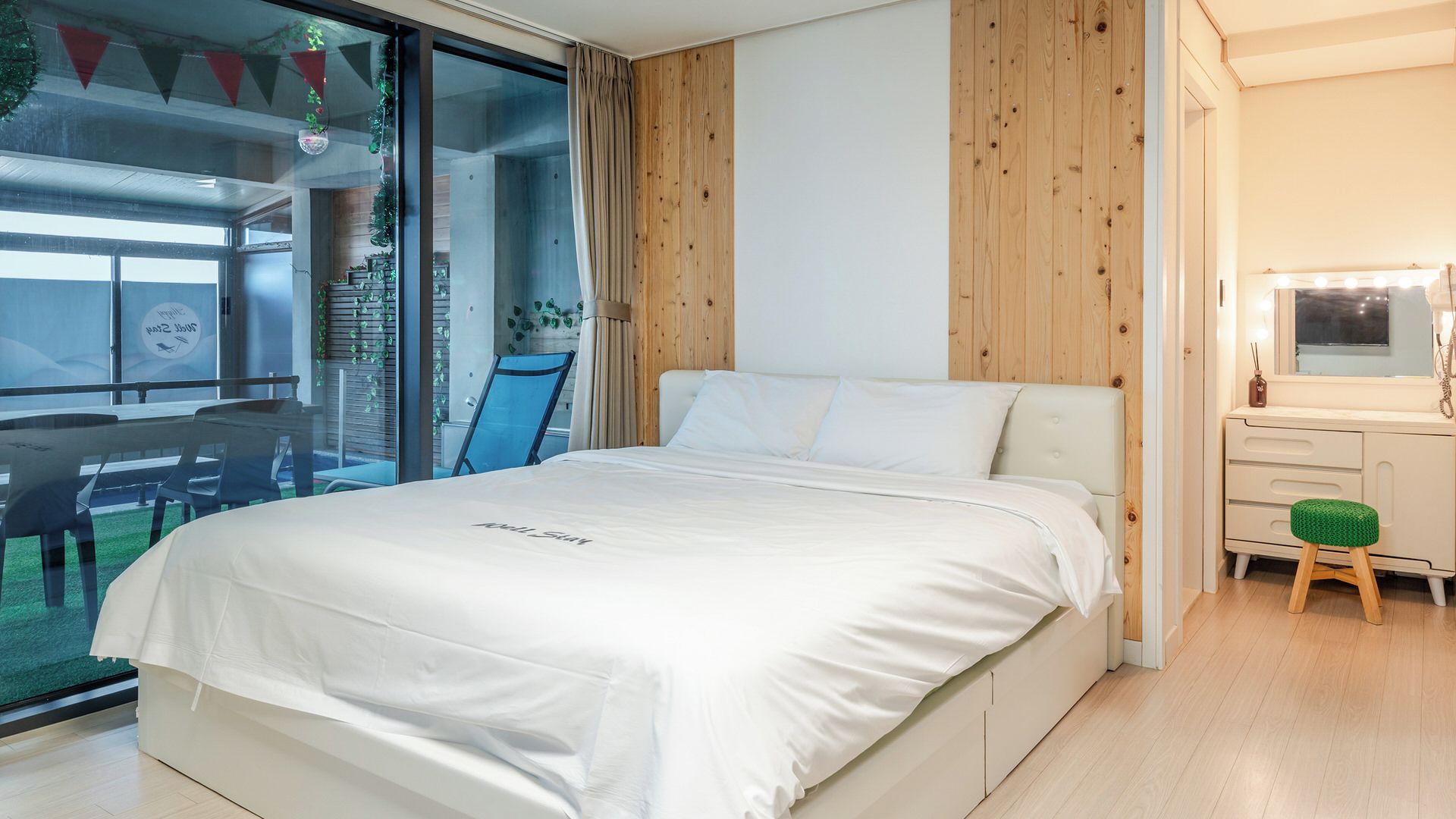 Quarto moderno com uma cama grande, painel de parede em madeira, um banquinho pequeno e uma toucador com iluminação.