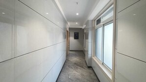 Um corredor com paredes de azulejos brancos e chão cinzento, que leva a um quarto com janela.