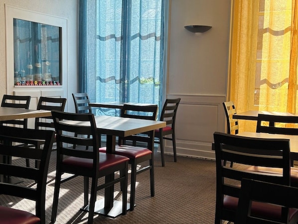 Uma sala de jantar com várias mesas e cadeiras, janelas amplas com cortinas azuis e vista para o exterior.