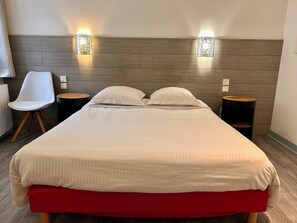 Cama bem arrumada com roupa de cama branca e saia vermelha, duas mesas de cabeceira com luminárias, uma cadeira branca e cabeceira de madeira.
