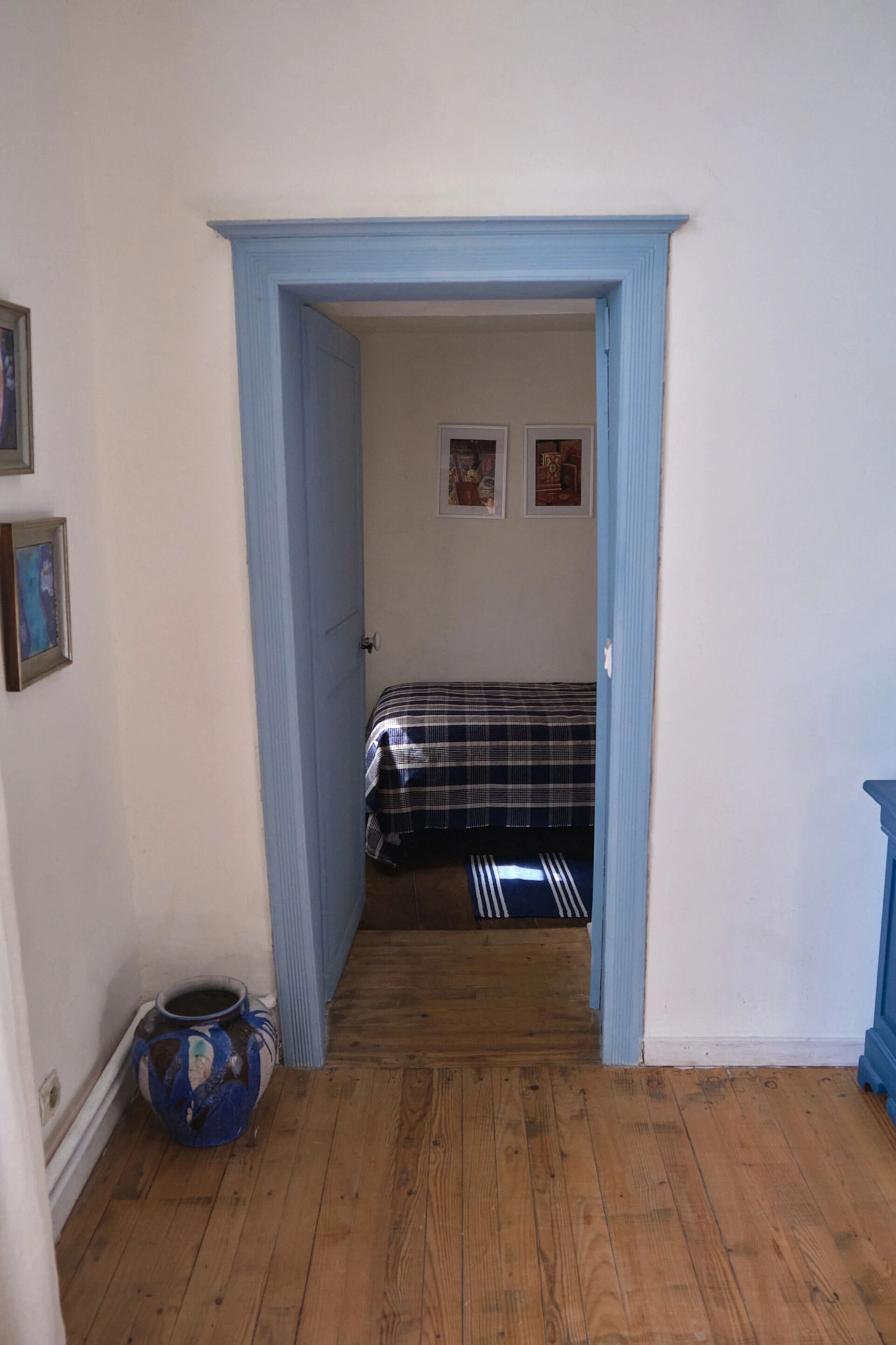Corredor que dá para um quarto com uma cama, porta azul e piso de madeira.