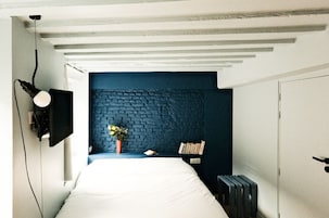 Quarto com cama branca, parede azul texturizada, um pequeno vaso com planta e uma estante de livros.