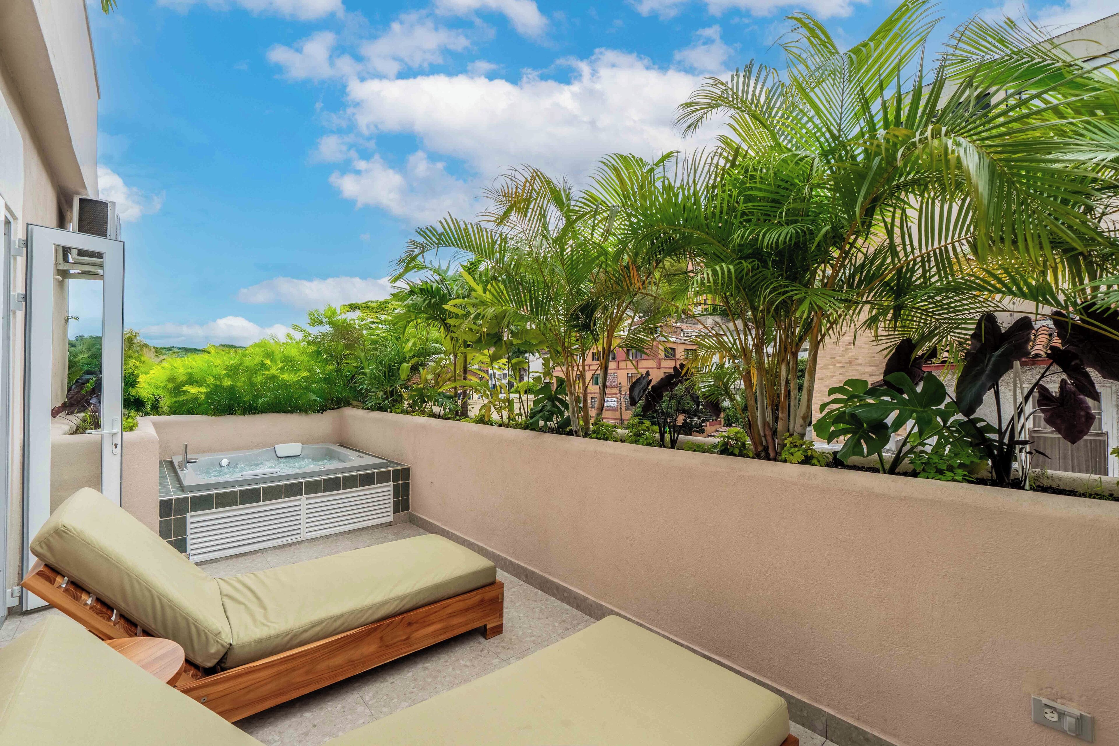 Um terraço no telhado com duas espreguiçadeiras de madeira, uma banheira de hidromassagem e plantas tropicais exuberantes.