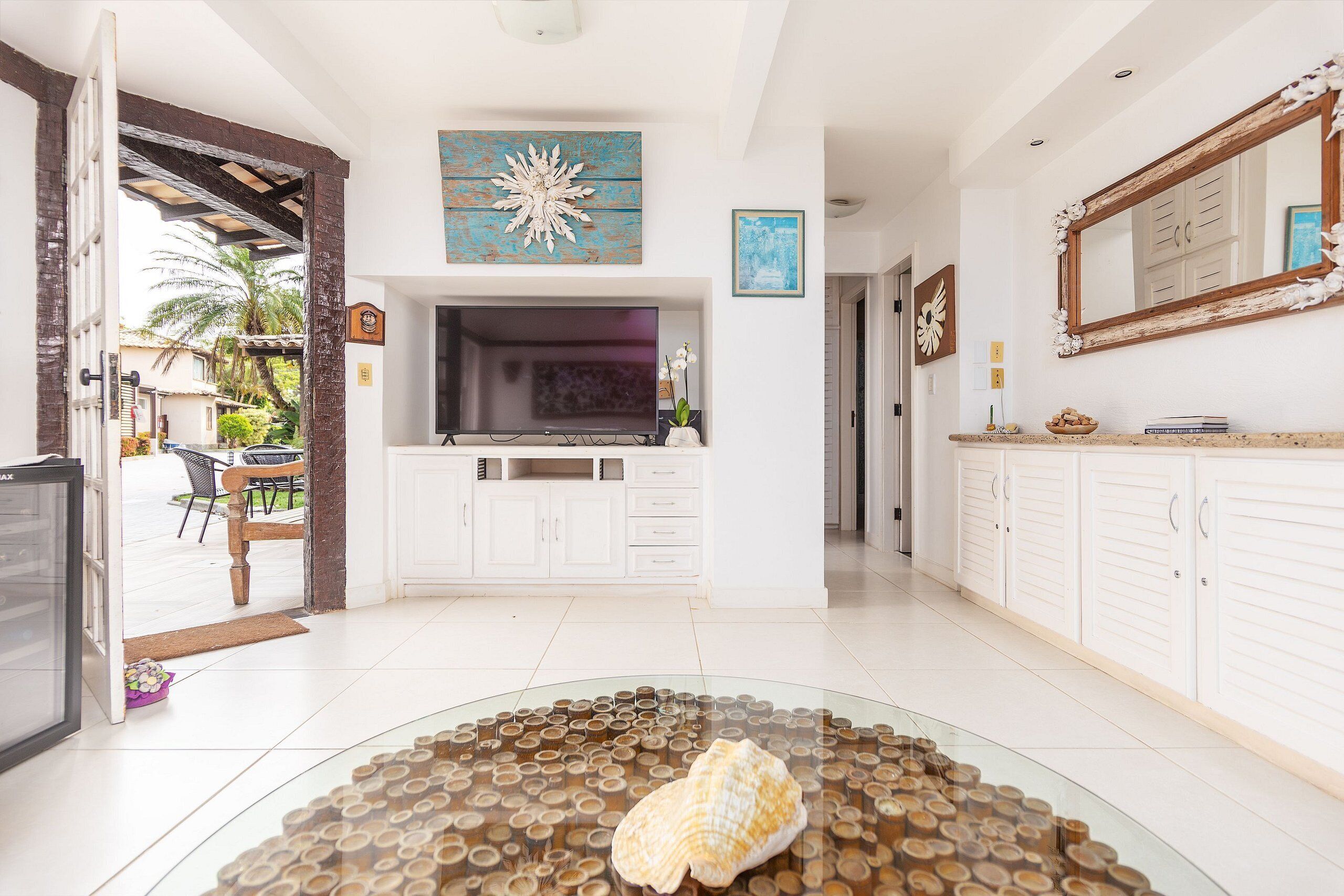 Uma sala de estar ampla com uma televisão grande, armários brancos e uma mesa de centro de vidro com um objeto decorativo.