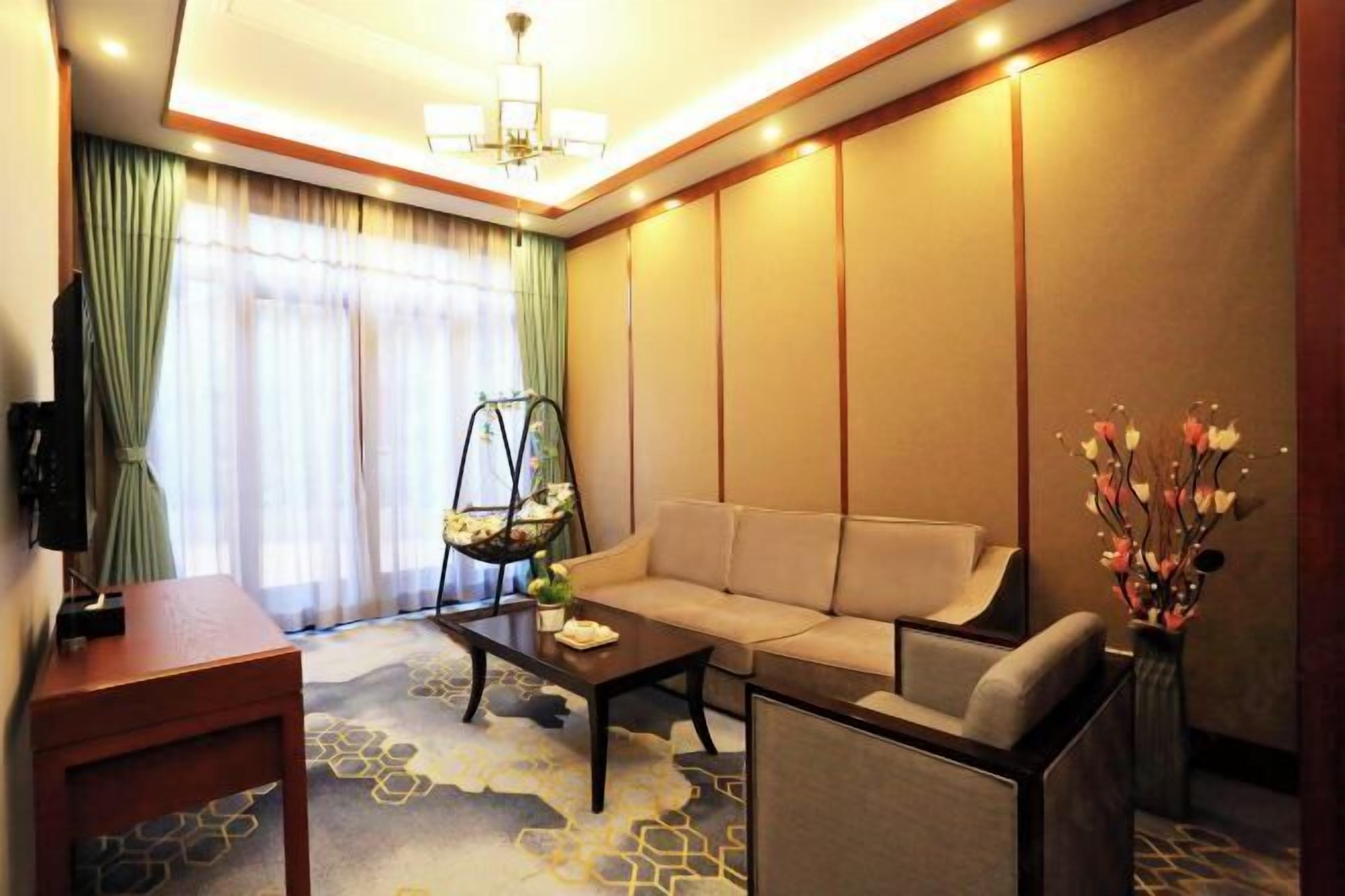 Quarto de hotel com sofá, mesa de centro, televisão, planta e janela com cortinas.