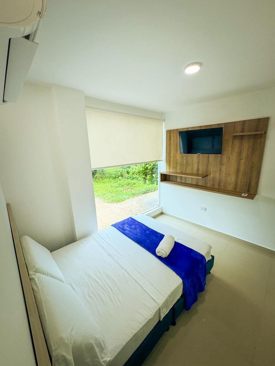 Quarto com cama, TV fixada na parede e janela com vista para área verde.