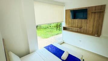 Quarto com cama, TV fixada na parede e janela com vista para área verde.