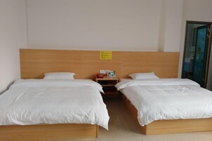 Quarto de hotel com duas camas de solteiro, cabeceira de madeira, mesa de cabeceira e uma pequena mesa com um livro e um objeto vermelho.