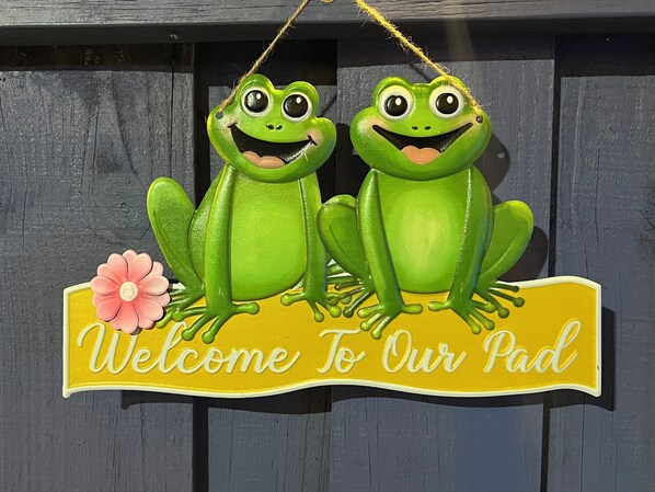 Um letreiro de madeira com duas decorações de sapo verde e uma flor rosa, com o texto 'Welcome To Our Pad'.